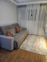Uma sala de estar moderna com um sofá cinzento, duas almofadas laranjas, um tapete fofinho e cortinas leves com padrão floral.