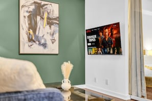 Quarto com pintura abstrata emoldurada, tela de Netflix na parede e uma pequena mesa com um vaso decorativo.