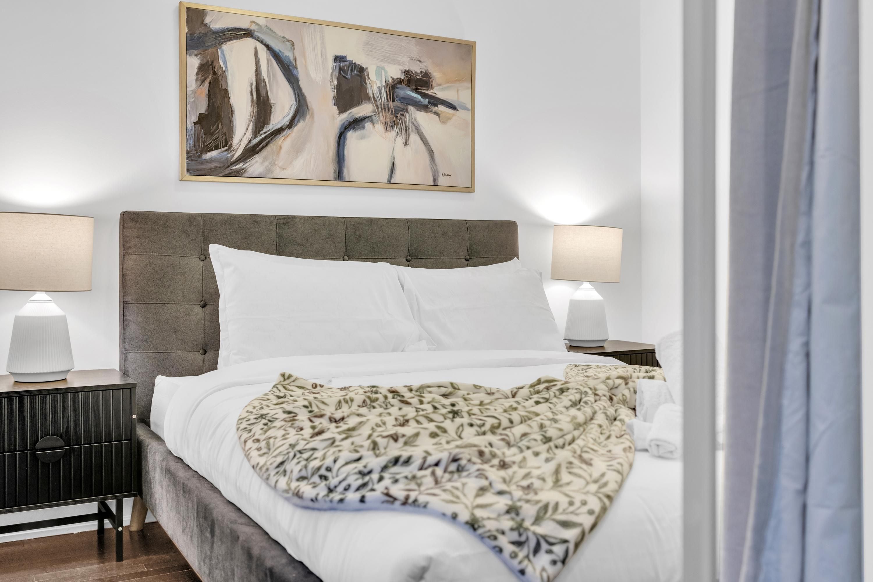 Quarto de hotel com cama grande, duas luminárias de cabeceira, um quadro decorativo e manta estampada.