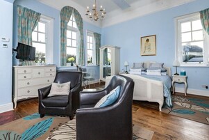 Quarto com cama, duas poltronas, cômoda, televisão e janela com cortinas.