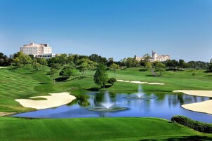 Um campo de golfe com bunkers, um lago com fontes e edifícios ao fundo.