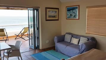 Quarto com sofá, mesa, cadeira, varanda com vista para o mar e duas quadros pendurados na parede.