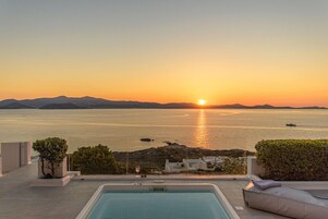 Área da piscina com vista para o mar e montanhas ao longe, durante o pôr do sol.