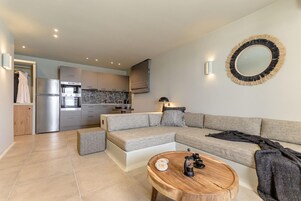 Uma sala de estar moderna com um sofá seccional, uma mesa de centro de madeira e uma cozinha ao fundo.
