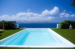Uma piscina retangular com água azul cristalina, rodeada por gramado bem cuidado, e com vista para um oceano extenso sob um céu claro.