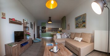 Uma sala de estar moderna com um sofá, mesa de centro e televisão. Cozinha ao fundo com armários verdes e uma área de jantar.