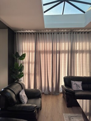 Sala de estar com claraboia, dois sofás de couro, uma planta grande e cortinas leves.
