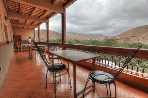 Um terraço ao ar livre com mesa e cadeiras de madeira, vista para uma paisagem desértica e céu nublado.