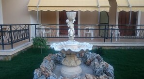 Uma fonte com uma estátua no centro, cercada por pedras e grama, com um edifício ao fundo.