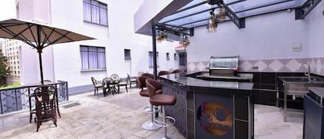 Área de bar ao ar livre com balcão, banquetas e estrutura de vidro.