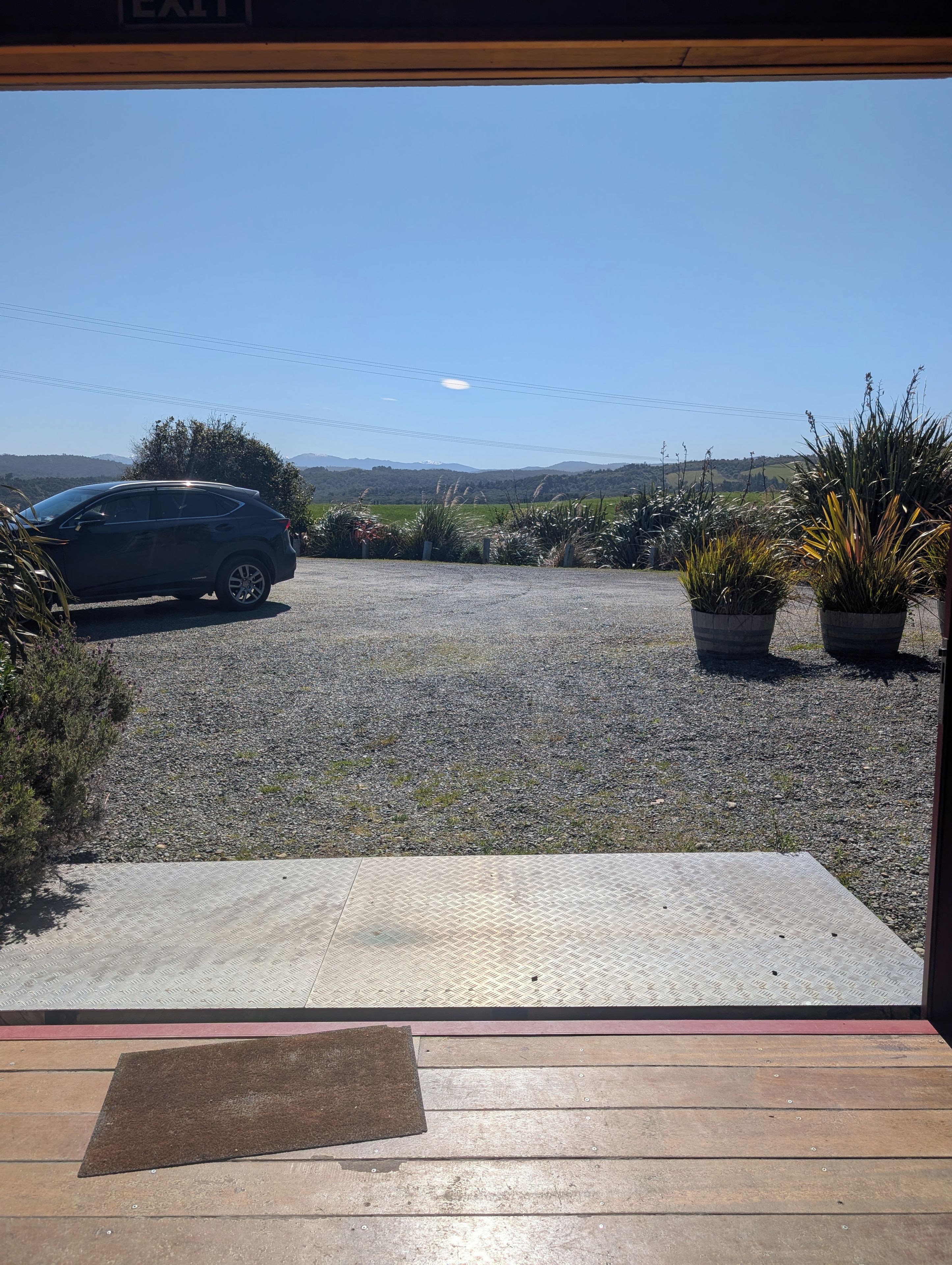 Vista do interior de um quarto para uma entrada de cascalho com um carro estacionado, plantas em vasos e céu claro.