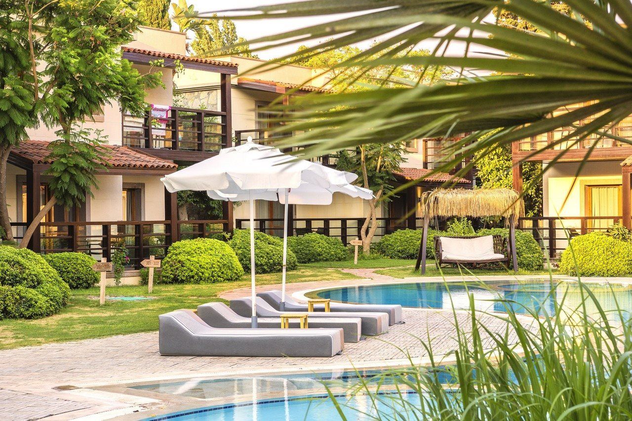 Área de piscina com espreguiçadeiras, um guarda-sol de palha e um edifício com telhado de azulejos.