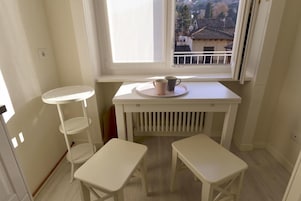 Um quarto pequeno e claro, com janela, radiador, uma mesa branca, duas cadeiras brancas e uma estante branca.
