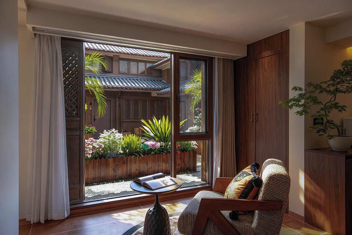 Quarto com porta de vidro deslizante que dá para um jardim, uma cadeira de madeira e uma pequena mesa com um livro.