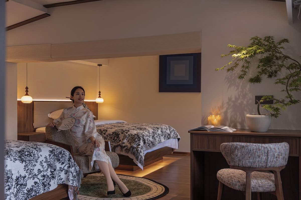 Quarto de hotel com duas camas, um espaço de estar com uma poltrona, mesa e planta em vaso.