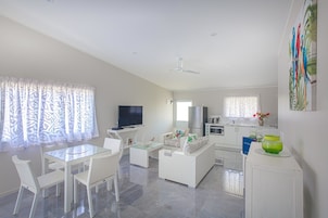 Uma sala de estar moderna com uma mesa de jantar de vidro, cadeiras de vime branco, uma TV de tela plana e uma área de cozinha com geladeira e micro-ondas.