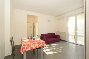 Uma sala de jantar com mesa posta para dois, um sofá vermelho e vista através de uma porta de vidro.