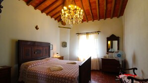 Quarto com teto de madeira, uma cama grande com colcha estampada, um lustre, uma mesinha de cabeceira com abajur, um espelho e um ventilador.