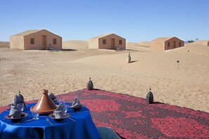 Cena de deserto com uma mesa posta para refeição, destacando uma toalha de mesa azul e um tapete vermelho com padrão.