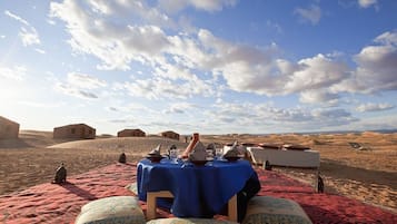 Paisagem desértica com mesa posta para refeição, cercada por almofadas e vista para dunas de areia sob céu parcialmente nublado.