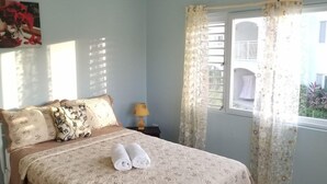 Quarto com cama, almofadas, abajur e janela com cortinas.