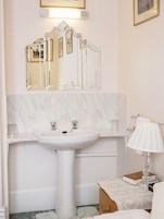 Um banheiro com pia de pedestal, espelho com moldura decorativa e mesa de cabeceira com abajur e um livro.