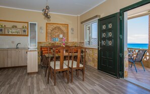 Uma sala de jantar com mesa e cadeiras, uma cozinha com pia e armários, e uma porta que dá para uma varanda com vista para o mar.