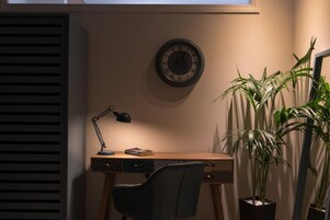 Uma mesa com um relógio, uma luminária, uma cadeira e um vaso de planta.