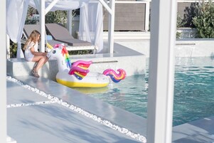 Área de piscina com um flutuador de unicórnio, uma criança sentada em uma espreguiçadeira e uma estrutura à beira da piscina.