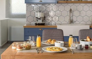 Mesa de madeira com máquina de café, diversos alimentos e acompanhamentos em cozinha moderna com armários azuis e painel de azulejos hexagonais.
