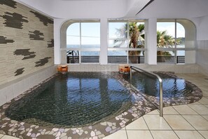 Área de spa com duas banheiras de hidromassagem, piso ladrilhado e amplas janelas com vista para um corpo de água.