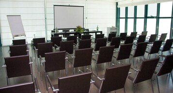 Uma sala de conferências com fileiras de cadeiras, uma tela de projetor e uma lousa.
