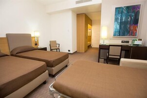 Quarto de hotel com duas camas de solteiro, uma escrivaninha com cadeira, luminária e um quadro na parede.
