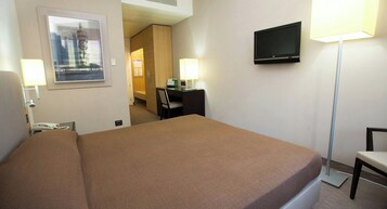 Quarto de hotel com cama, escrivaninha, cadeira, televisão e abajur.