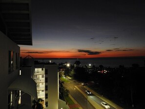 Vista noturna de uma varanda com vista para uma rua, prédios e um pôr do sol.