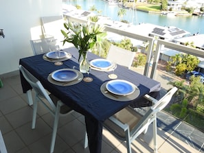 Mesa de jantar posta com pratos azuis, um vaso de flores e um copo de água numa varanda com vista para uma marina.