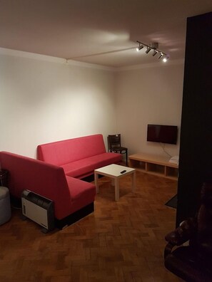Uma sala de estar com um sofá vermelho, uma mesa pequena branca, um banco de madeira, uma televisão e um lustre no teto.