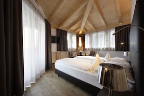 Quarto de hotel com teto de madeira, cama com roupa de cama branca, mesa de cabeceira com abajur e uma grande janela com cortinas.