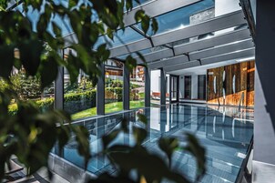 Piscina interior moderna com paredes de vidro, cercada por vegetação e vista para um edifício.