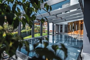 Piscina interior moderna com paredes de vidro, cercada por vegetação e vista para um edifício.