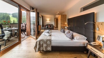 Quarto com cama grande, mesa de cabeceira com abajur, cabeceira de madeira e vista para uma varanda com mesa e cadeiras.