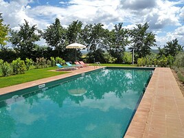 Piscina retangular com borda azulejada, rodeada por um deck de tijolos. Há espreguiçadeiras e uma área com sombra e um guarda-sol.
