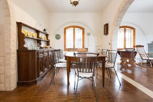 Uma sala de jantar com mesa redonda, cadeiras, um aparador de madeira com copos e uma grande janela com desenho em arco.