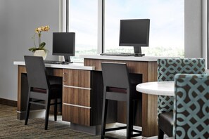 Uma mesa de escritório moderna com dois monitores, um teclado e uma planta em vaso. Há duas cadeiras e uma pequena mesa redonda com uma almofada estampada.
