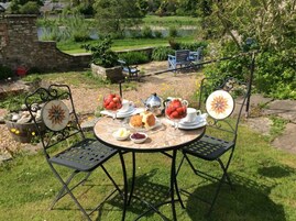 Uma área de refeições ao ar livre com mesa redonda e cadeiras, rodeada por um jardim com muro de pedra e vegetação.