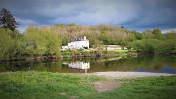 Um edifício branco com um lago à frente, rodeado por árvores e um céu nublado.