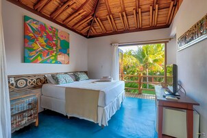 Quarto com cama, escrivaninha, televisão e varanda com vista para palmeiras.