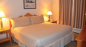 Uma cama bem arrumada com cabeceira, duas luminárias de apoio, um quadro na parede e uma janela com cortinas.