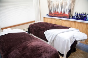 Quarto com duas camas cobertas por lençóis escuros e brancos, um armário de madeira com garrafas e uma pintura de uma floresta.