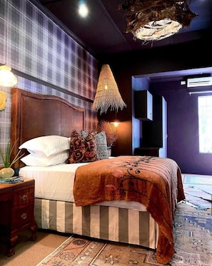 Quarto com cama grande, cabeceira de madeira, luminária trançada e tapete estampado.